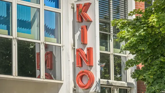 Ein altmodischer, vertikaler Schriftzug "Kino" an einer Fensterfront 