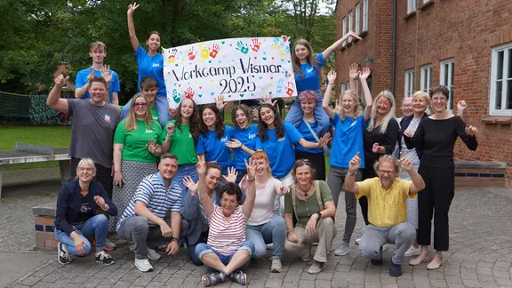Gruppenbild mit etwa 20 Menschen. Sie jubeln in die Kamera und ein Plakat mit der Aufschrift "Workcamp Wismar 2025" ist zu sehen.