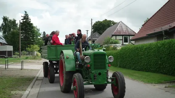 Auf einer Dorfstraße: Ein grüner Oldtimer-Traktor mit Anhänger, auf dem mehrere Personen sitzen.